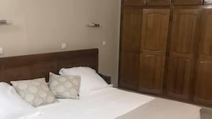 Uma cama bem arrumada com lençóis brancos e almofadas estampadas, um guarda-roupa de madeira e um chão de azulejos.