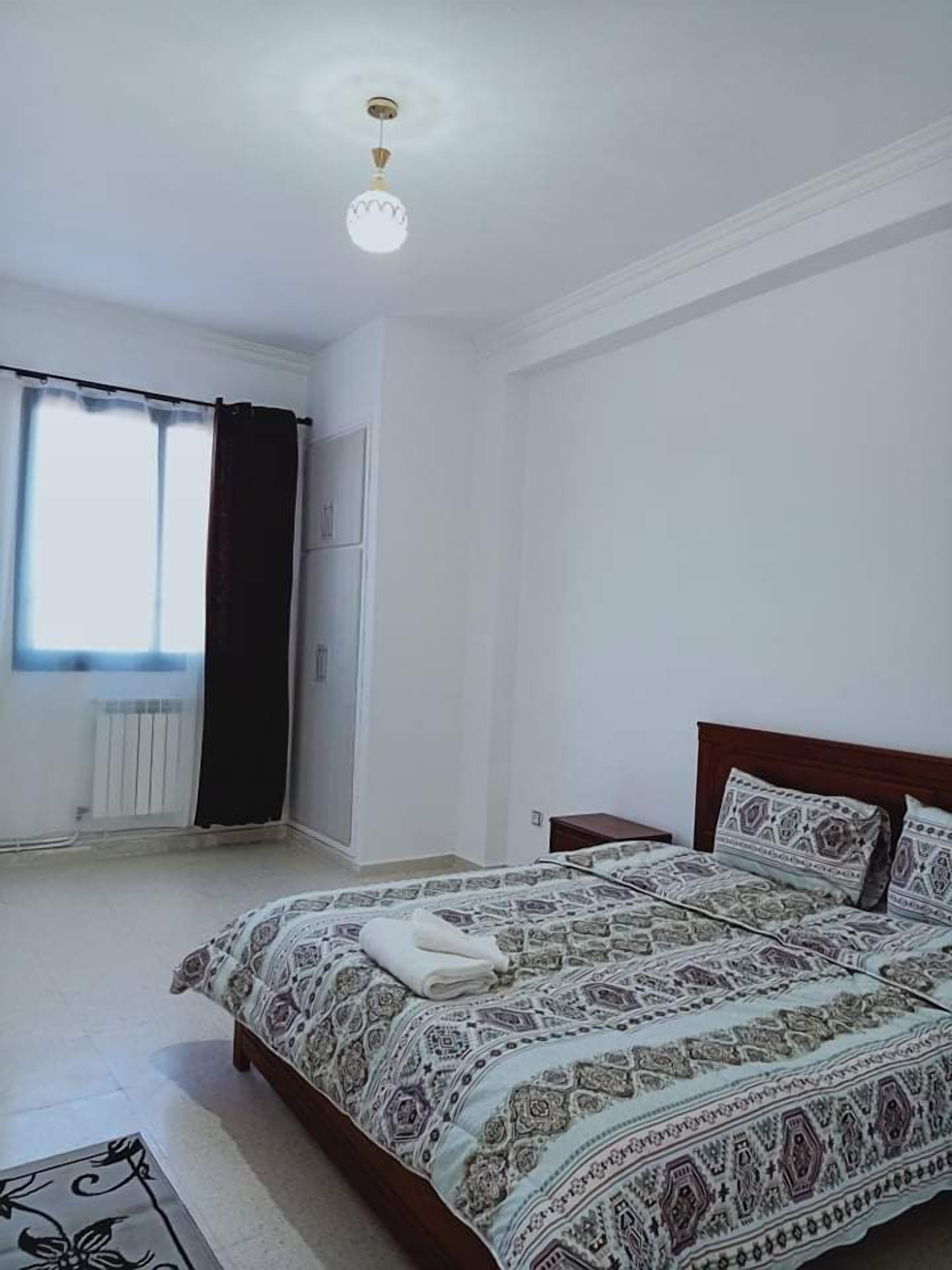 Quarto com cama, mesa de cabeceira, janela com cortinas e radiador.
