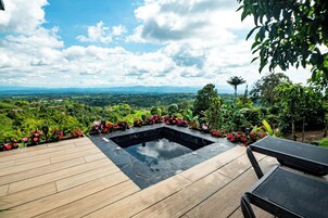 Um deck de madeira com uma banheira de hidromassagem retangular, rodeada por flores vibrantes, com vista para uma paisagem exuberante e montanhas ao longe.