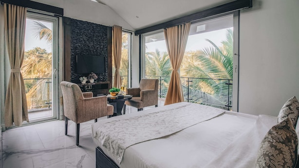 Quarto de hotel moderno com uma cama grande, duas poltronas, uma pequena mesa com uma planta, televisão de tela plana e varanda com vista para palmeiras.