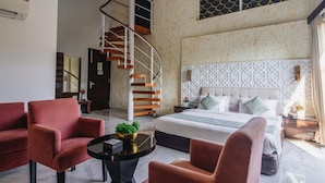 Quarto de hotel moderno com uma cama grande, escada de madeira, poltronas vermelhas e uma pequena mesa redonda com uma planta.