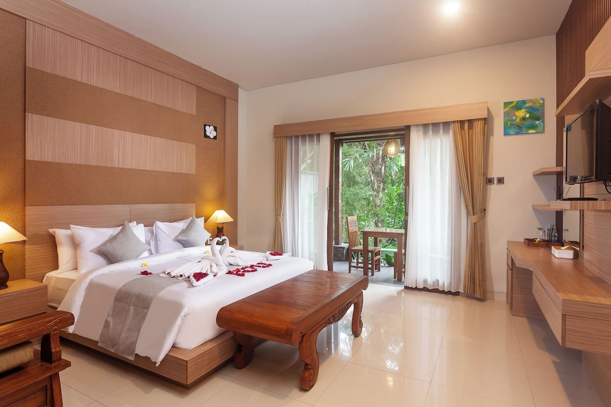 Quarto de hotel com uma cama grande, mesas de cabeceira, um banco de madeira, televisão e uma varanda com cortinas.