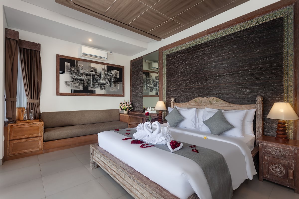 Quarto de hotel com uma cama grande, um sofá, uma cabeceira de madeira, um espelho e um quadro emoldurado na parede.