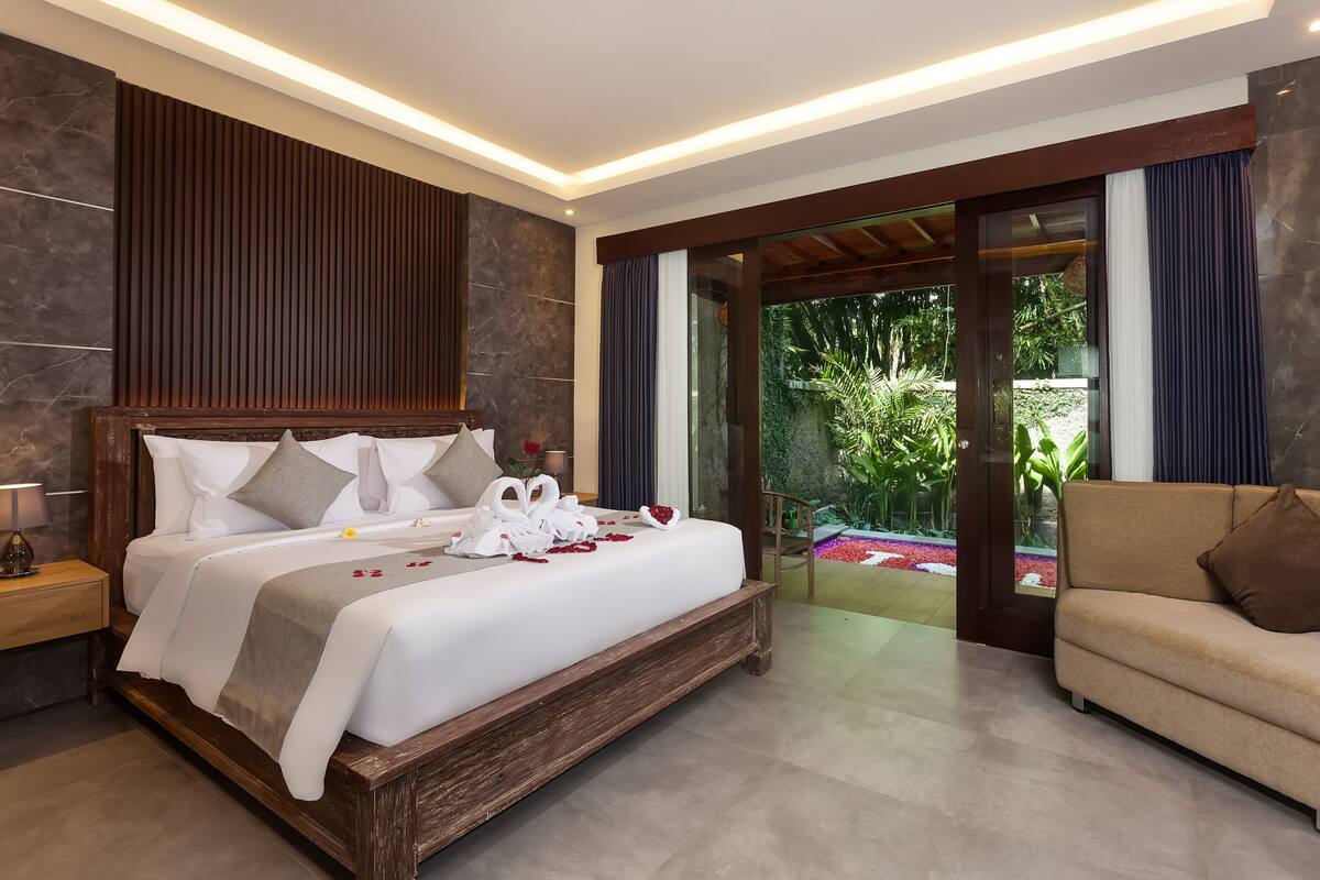Quarto de hotel moderno com uma cama grande, um sofá e vista para um jardim através de uma porta de vidro.