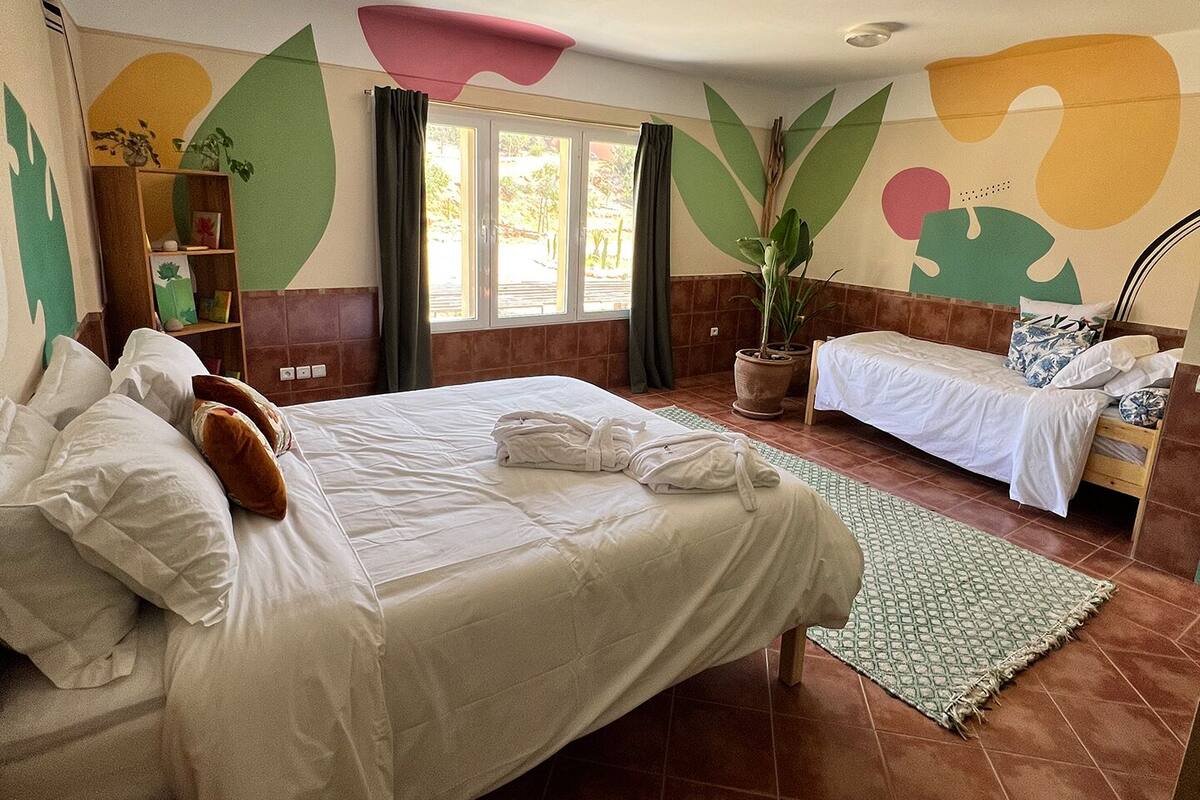 Quarto com uma cama grande, uma cama menor, um vaso de planta e uma parede com arte abstrata.