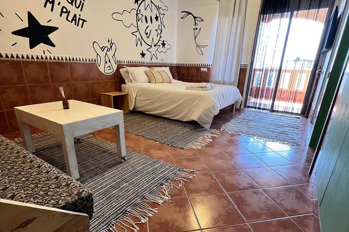 Quarto de hotel com uma cama, uma mesinha, um painel decorativo e vista pela janela.