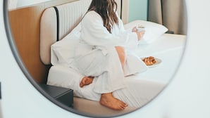 Uma pessoa sentada em uma cama em um quarto, com um espelho redondo refletindo parte do ambiente.
