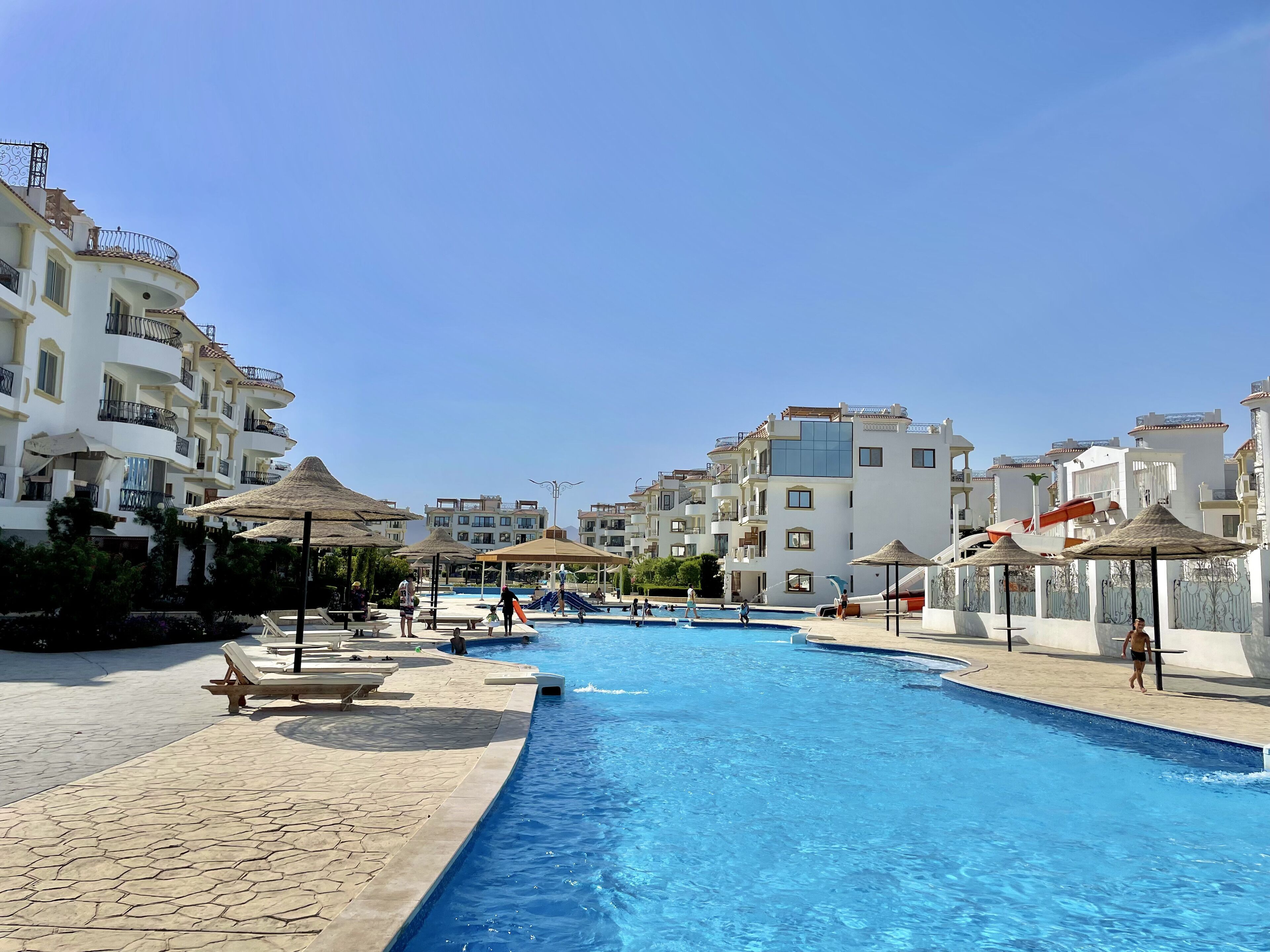 Uma área de piscina com espreguiçadeiras e guarda-sóis, cercada por edifícios com várias andares e varandas.