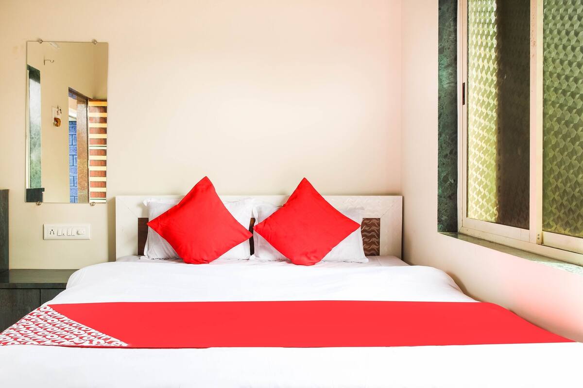 Quarto de hotel com cama, duas almofadas vermelhas, um edredom branco e uma janela com persiana texturizada.