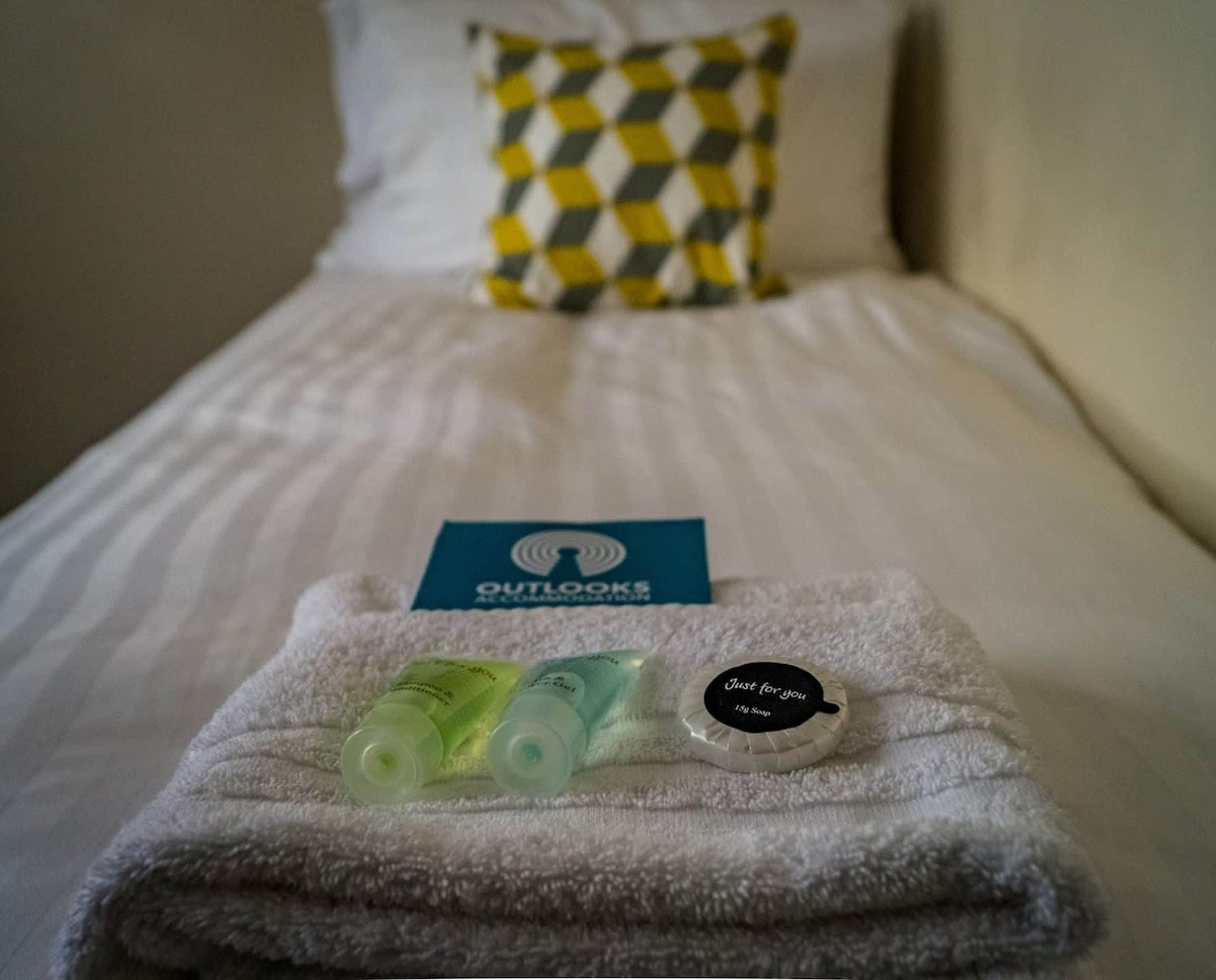 Cama bem arrumada com travesseiro quadriculado branco e amarelo, toalha dobrada e pequeno cartão azul com o logo 'OUTLOOK' em um quarto de hotel.