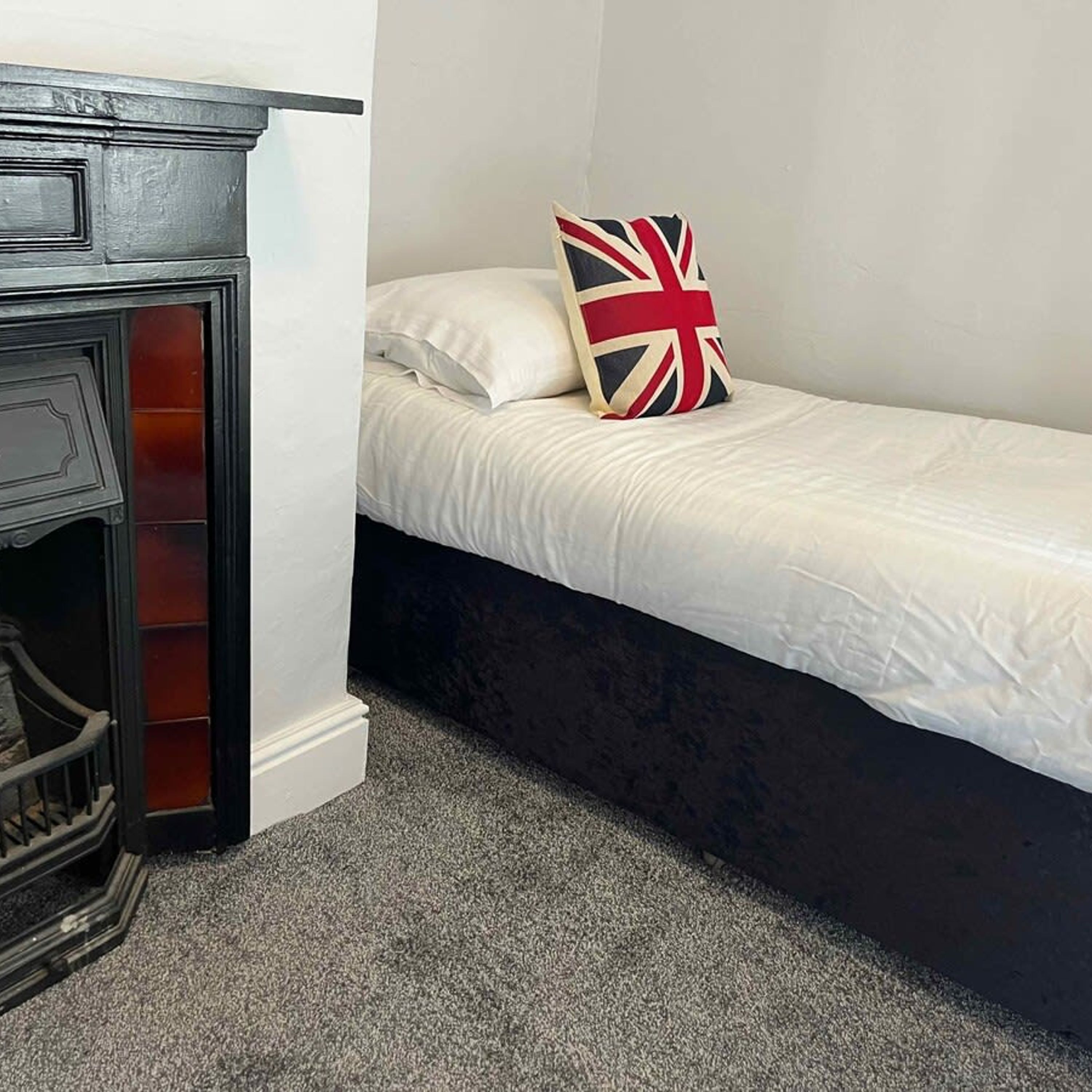 Quarto com cama, travesseiro com estampa da Union Jack, cabeceira escura e piso coberto de carpete.