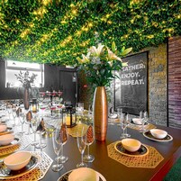 Um restaurante com teto coberto de folhagem verde, um vaso grande com flores e uma placa que diz 'GATHER. ENJOY. REPEAT'.
