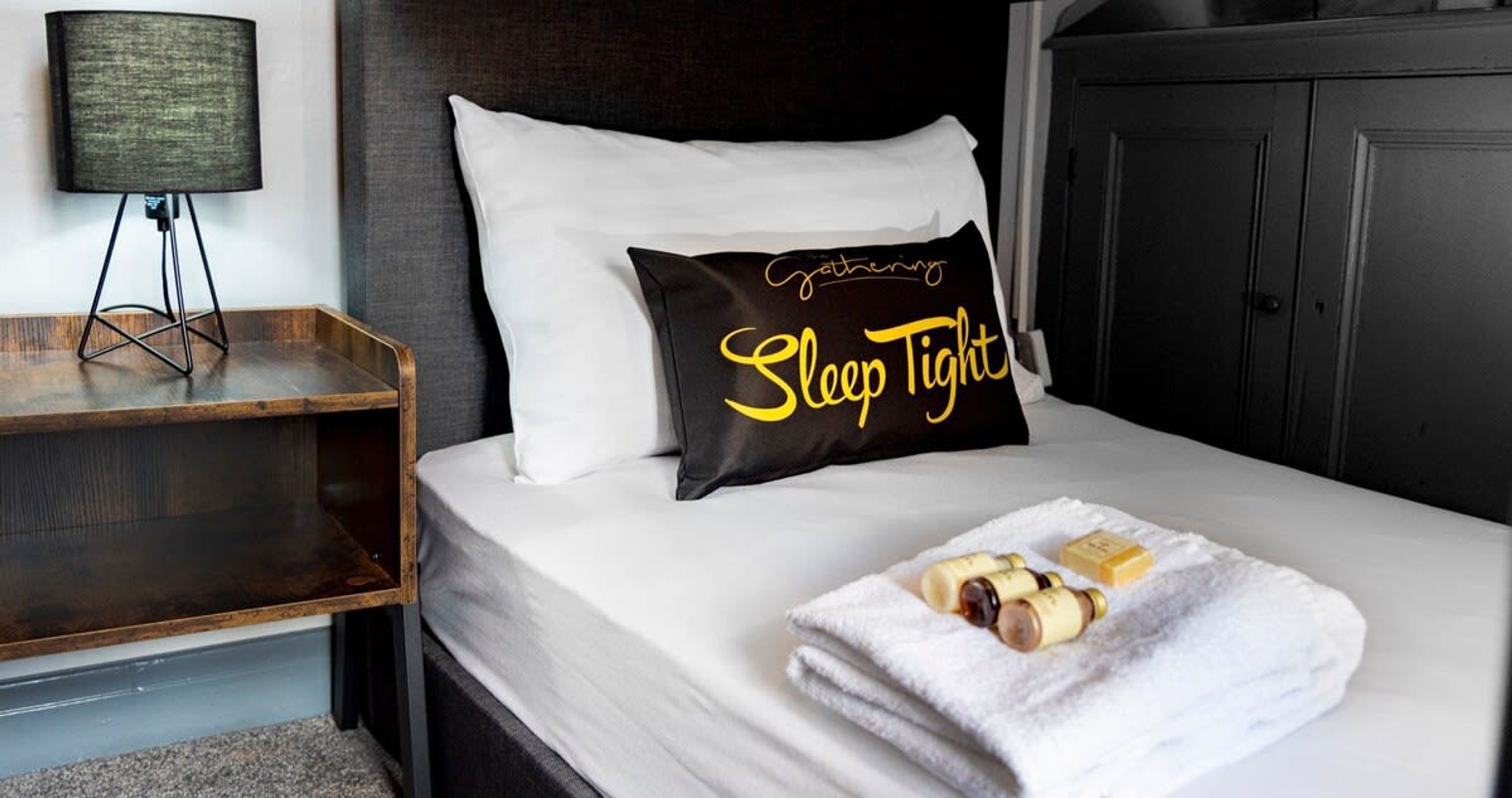 Cama bem arrumada com travesseiro que diz 'Gathering Sleep Tight' e mesa de cabeceira com luminária e artigos de higiene pessoal.