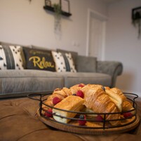 Uma bandeja com croissants e frutas vermelhas sobre uma mesa marrom em uma sala de estar com um sofá cinza e uma almofada com a palavra 'Relax'.