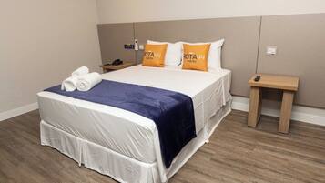 Quarto de hotel com uma cama bem arrumada, uma mesa de cabeceira com telefone e uma pequena mesinha de madeira.