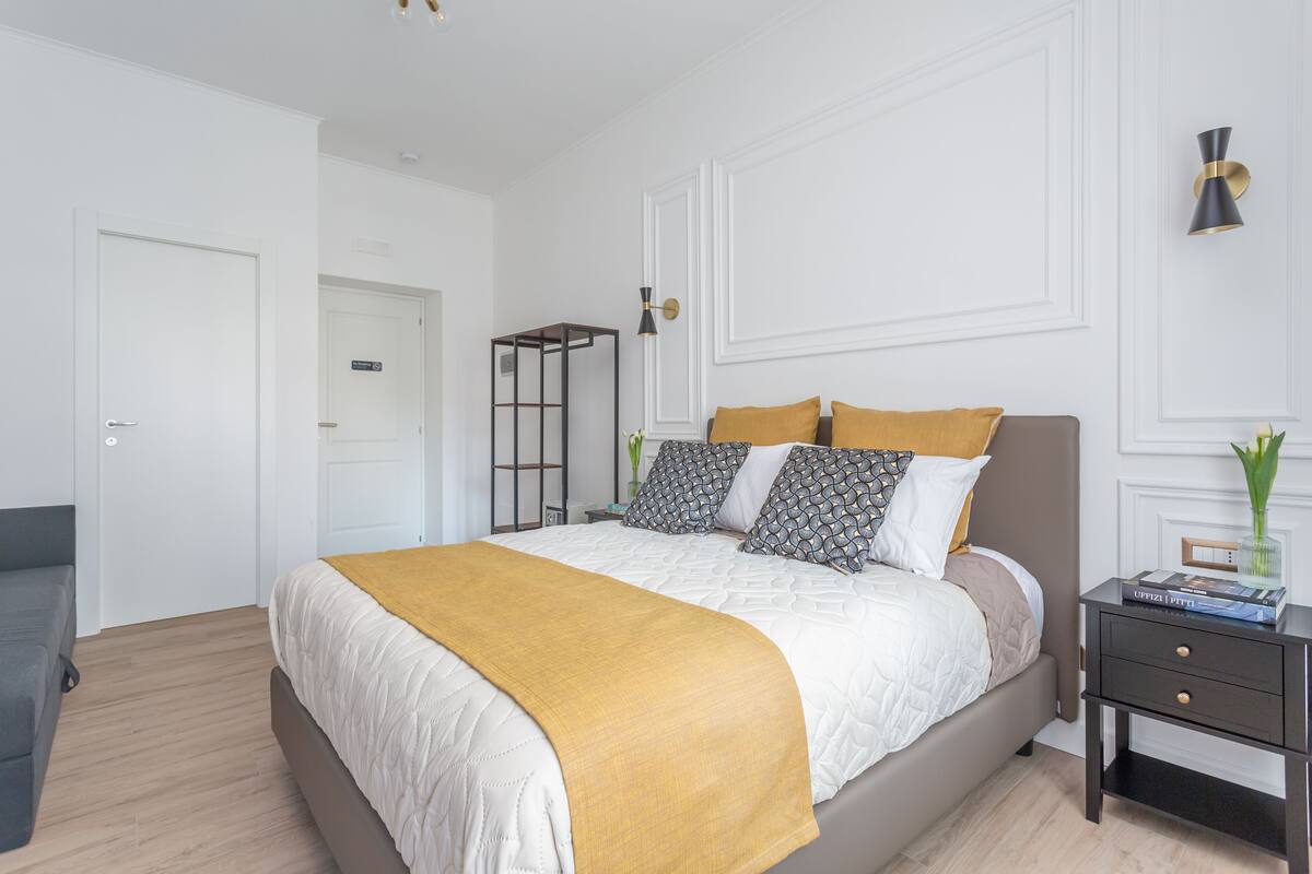 Quarto moderno com uma cama grande, mesa de cabeceira com abajur e porta ao fundo.