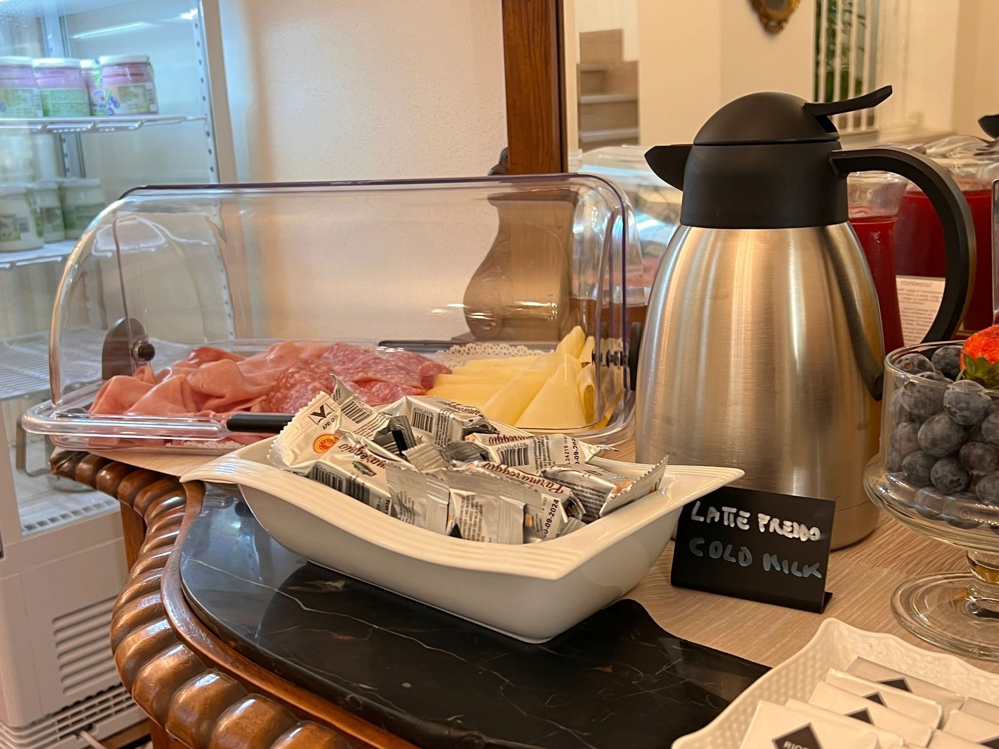 Disposição de buffet com diversos alimentos, um jarro de café e uma placa com a inscrição 'LATTE FREID COLO NICK'.