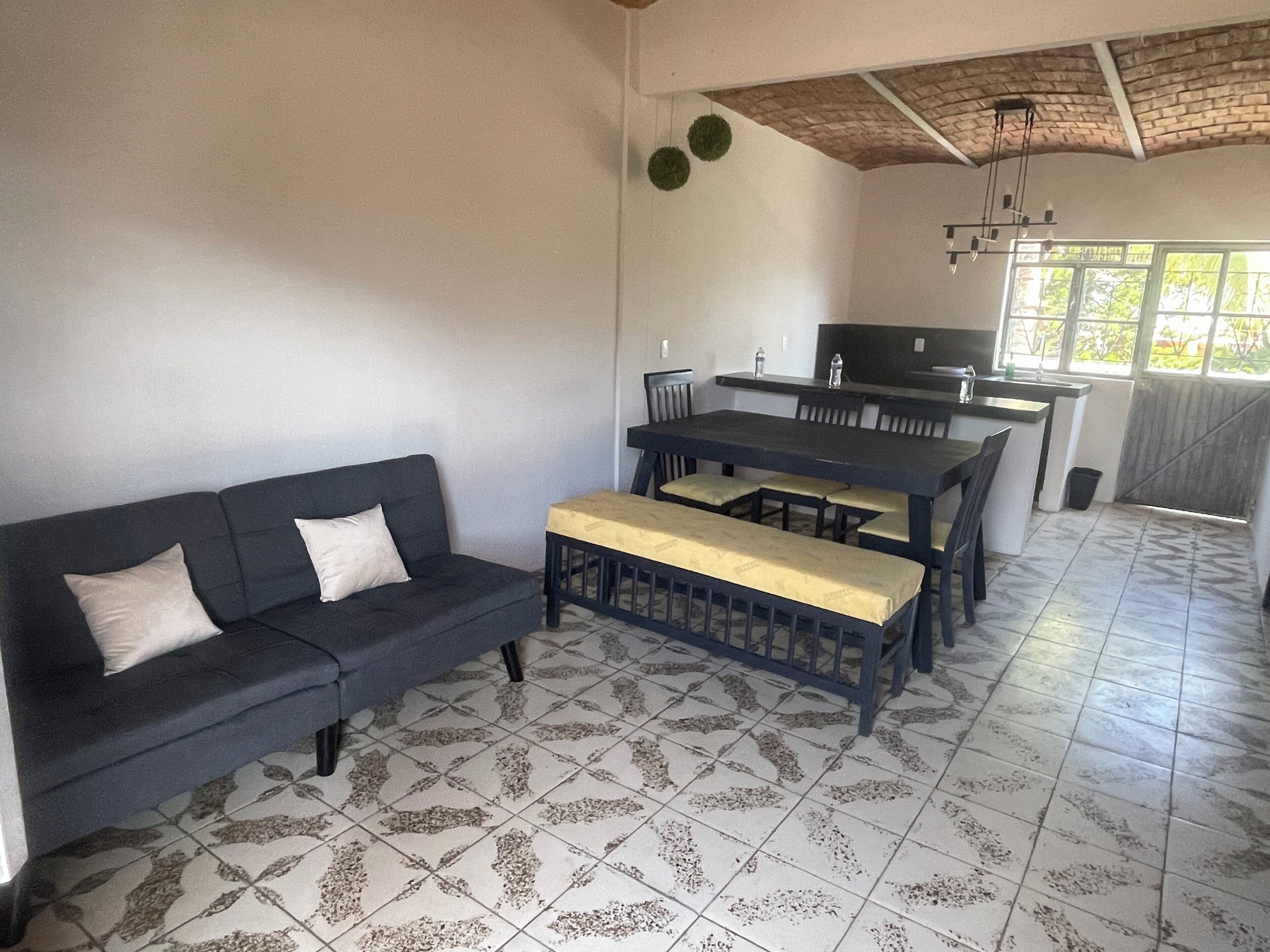 Uma área de refeições com um sofá de cor escura, um banco e uma mesa com cadeiras. Há também uma cozinha com uma pia e uma janela.