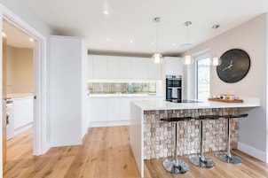 Cozinha moderna com armários brancos, uma ilha central com banquetas, iluminação pendente e um relógio redondo grande na parede.