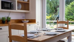 Uma sala de jantar com mesa de madeira posta para quatro, armários brancos, micro-ondas e vista para o verde lá fora.