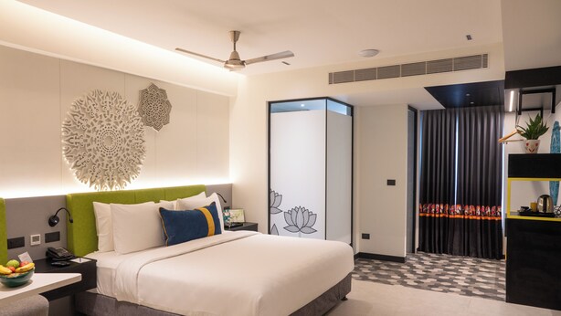 Quarto de hotel moderno com uma cama grande, uma escrivaninha com uma cesta de frutas, uma mesinha de cabeceira, um ventilador de teto, painéis decorativos na parede e vista para o exterior através de uma janela com cortinas.