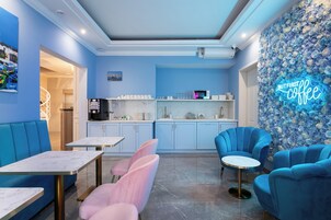 Um interior moderno com paredes azuis, um letreiro de neon que diz "BUTFIRST coffee" e áreas de estar com cadeiras cor de rosa e azuis.