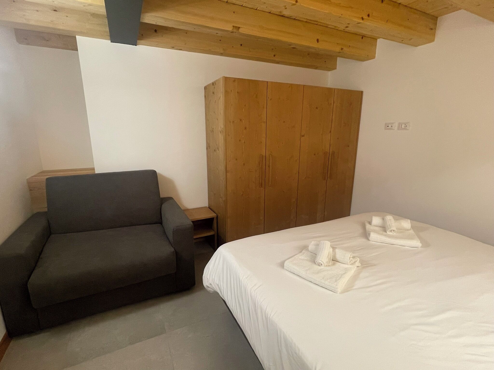 Um quarto com uma cama, um sofá cinzento, um guarda-roupa de madeira e uma pequena mesa de madeira.