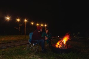 Duas pessoas sentadas perto de uma fogueira à noite, com postes de luz ao fundo.