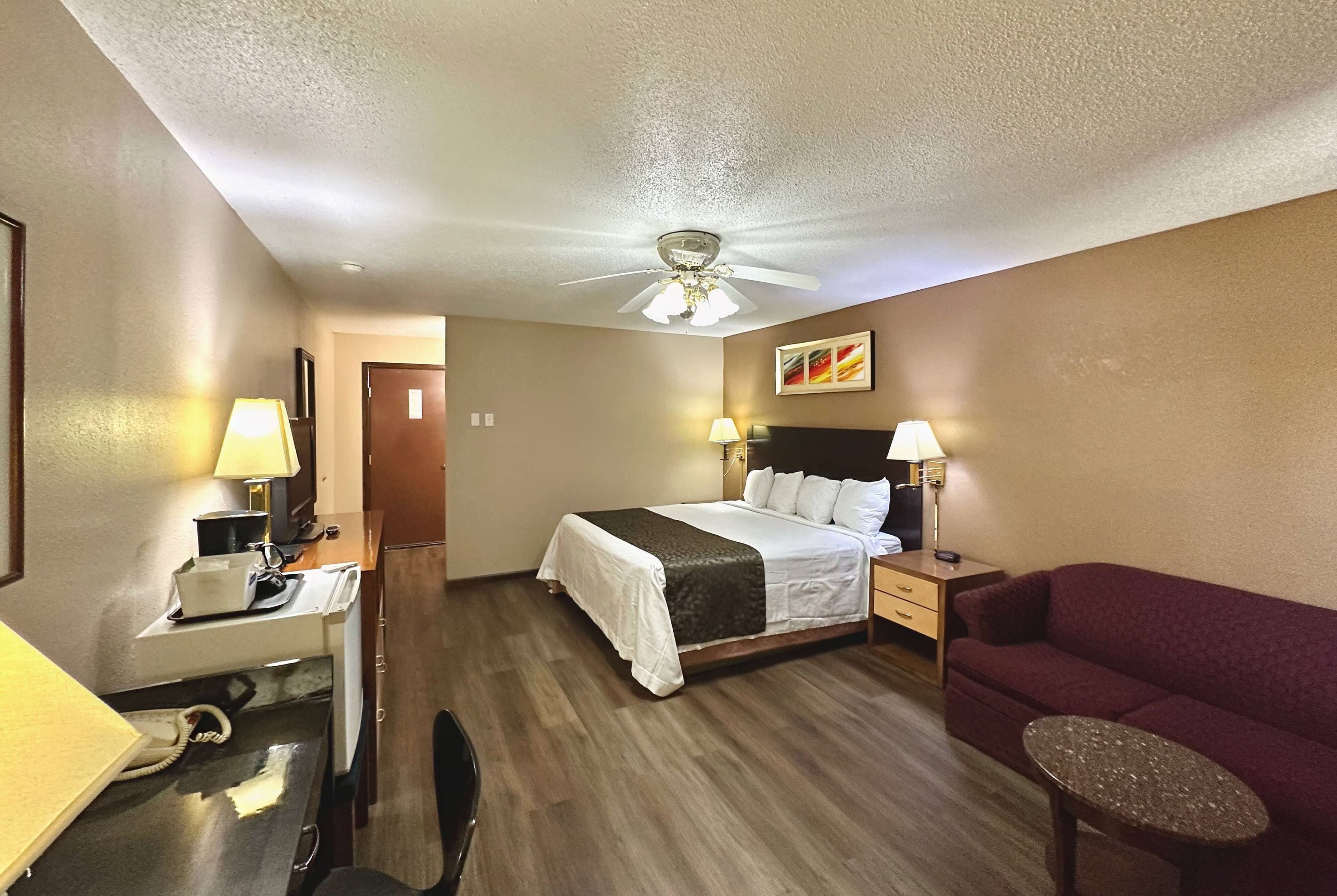 Quarto de hotel com cama, mesas de cabeceira com luminárias, escrivaninha com telefone, cadeira, sofá vermelho e ventoinha no teto.