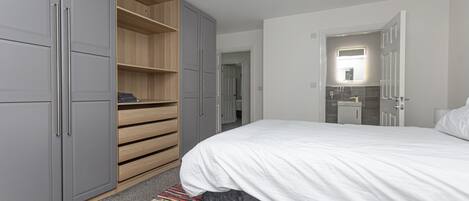 Quarto com cama grande, guarda-roupa cinzento, estantes de madeira e banheiro visível através de uma porta aberta.