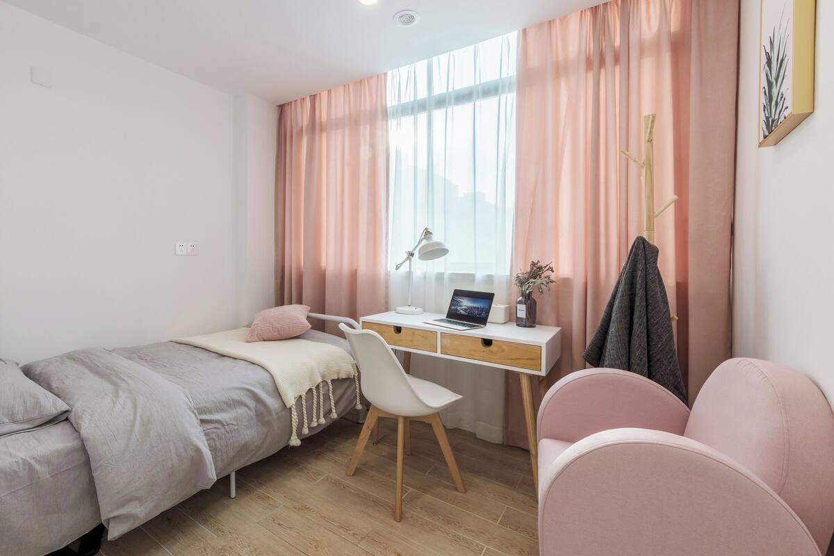Quarto com cama, escrivaninha, cadeira e janela com cortinas.