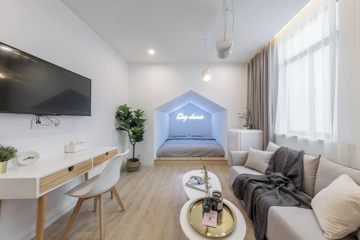 Quarto moderno e bem iluminado, com televisão de tela plana, escrivaninha com cadeira, cama com placa 'Day dream', sofá e mesa de centro com bandeja.