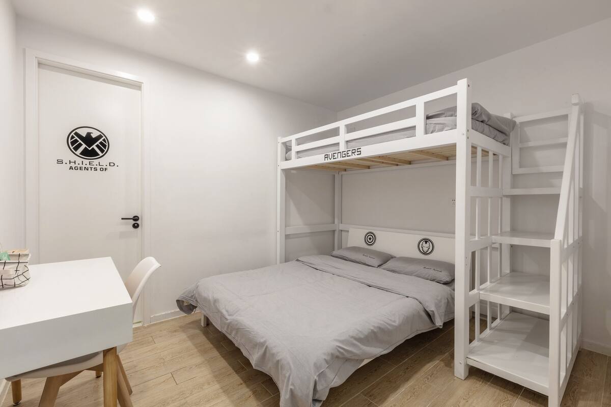 Quarto com uma cama beliche identificada com a inscrição 'AVENGERS', uma escrivaninha branca e uma porta com um logotipo.