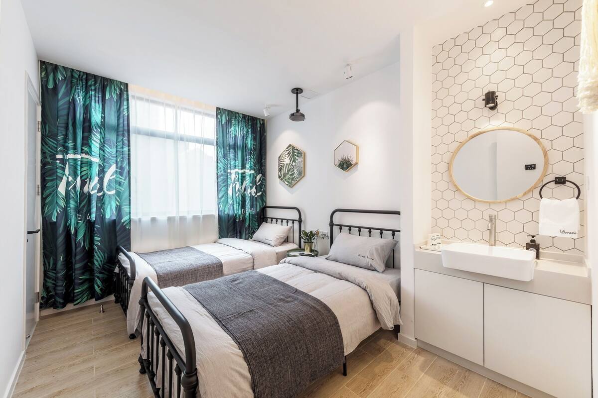 Quarto com duas camas, um espelho redondo, painel de azulejos hexagonais e uma janela com cortinas.