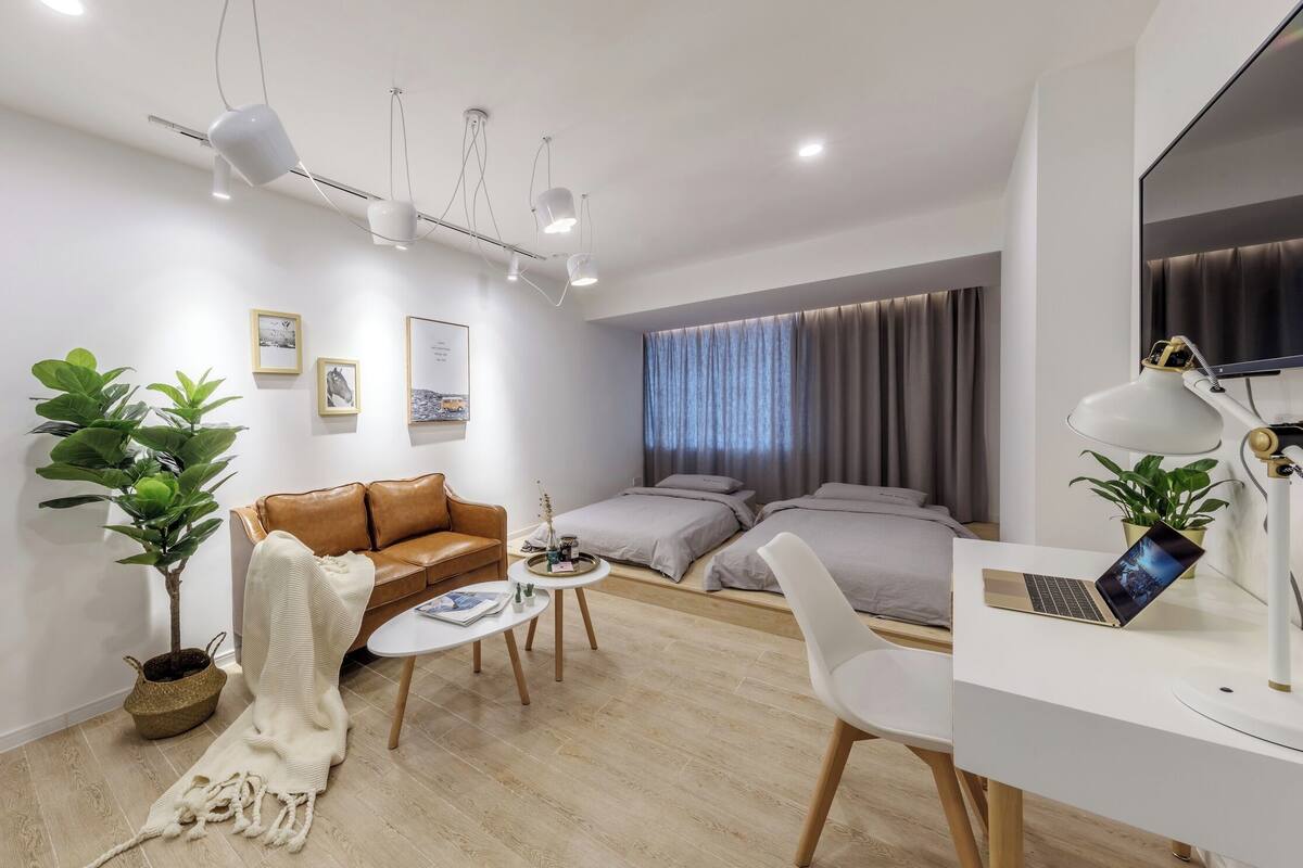 Quarto de hotel moderno com sofá de couro marrom, escrivaninha branca com um laptop, uma mesa redonda pequena e uma cama com roupa de cama cinza. Há duas plantas em vasos, um cobertor branco e uma TV fixada na parede.