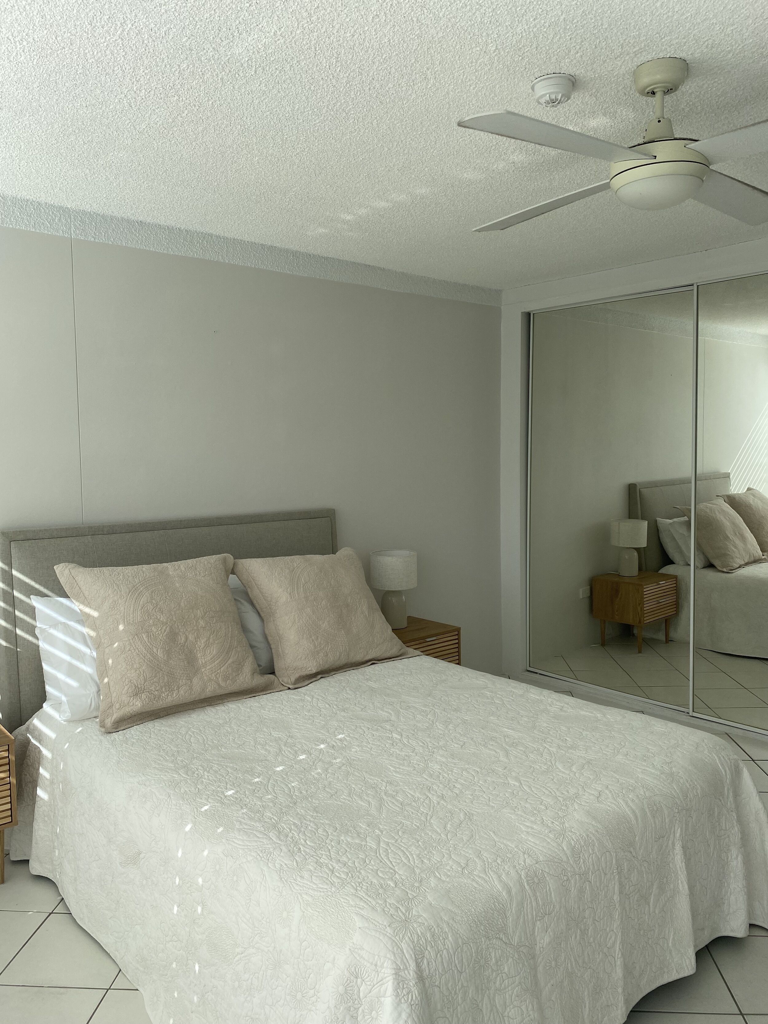 Quarto com cama, ventoinha no teto, mesa de cabeceira, abajur e espelho.