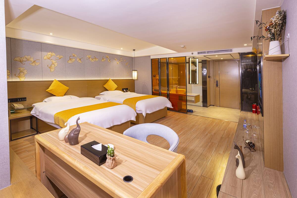Quarto de hotel com duas camas, uma mesa, uma cadeira, um guarda-roupa e um banheiro visível através de uma porta de vidro.