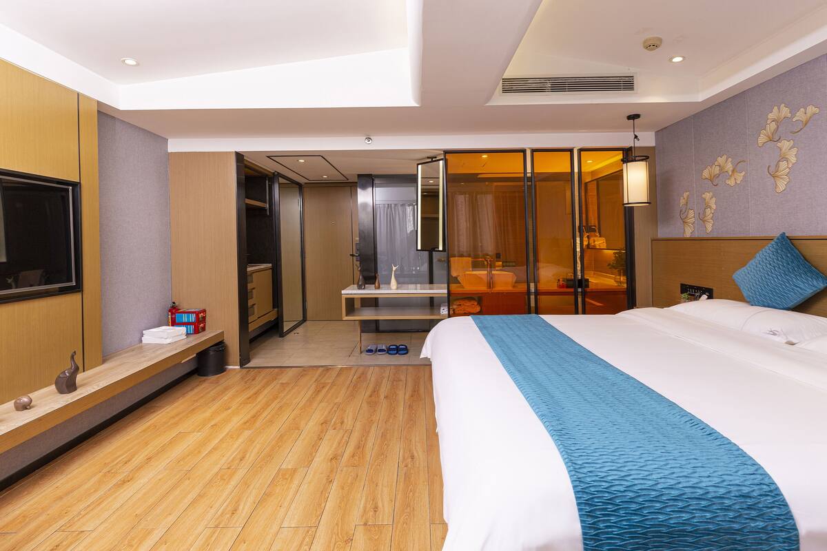 Quarto de hotel com uma cama grande, televisão, banheiro com espelho e piso de madeira.