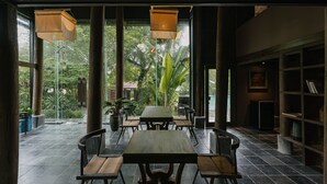 Amplo espaço interno com uma mesa grande e cadeiras, cercado por janelas altas e plantas em vasos, oferecendo vista de vegetação exuberante.