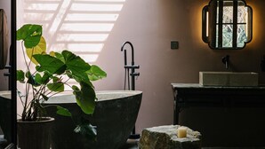 Casa de banho com banheira de apoio, lavatório, planta em vaso e detalhe em pedra.