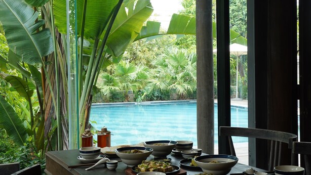 Uma área de refeições com vista para uma piscina, rodeada por grandes folhas de bananeira e plantas tropicais.