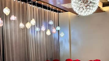 Uma sala de banquetes com mesas redondas cobertas por toalhas brancas, cadeiras vermelhas e um lustre decorativo.