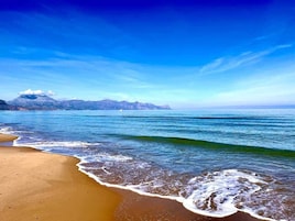 Uma praia com águas azul-claras, ondas e montanhas ao longe sob um céu azul.