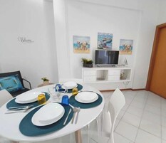 Uma sala de jantar com uma mesa redonda posta para quatro pessoas, cadeiras brancas e um armário branco com uma TV e objetos decorativos.