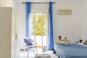 Quarto com cama, cadeira, mesa pequena com xícara e flores, e janela com cortinas.