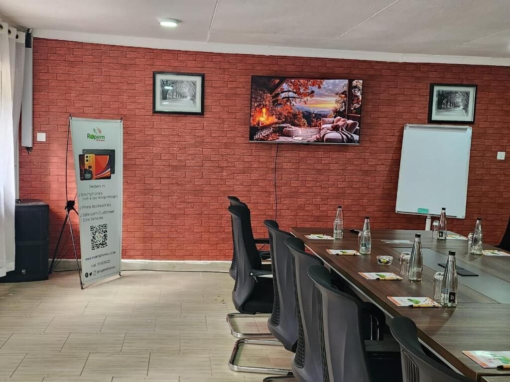 Uma sala de conferências com parede de tijolos, uma grande tela de TV, um quadro branco e uma mesa com cadeiras e garrafas de água.