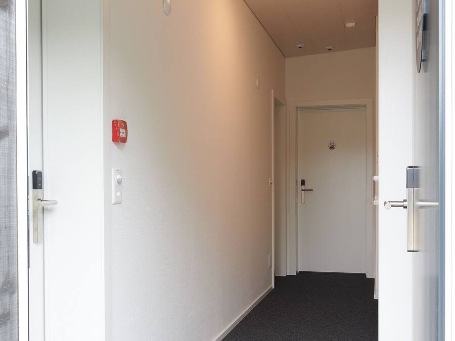 Um corredor com paredes brancas, carpete cinzento e uma porta no final.