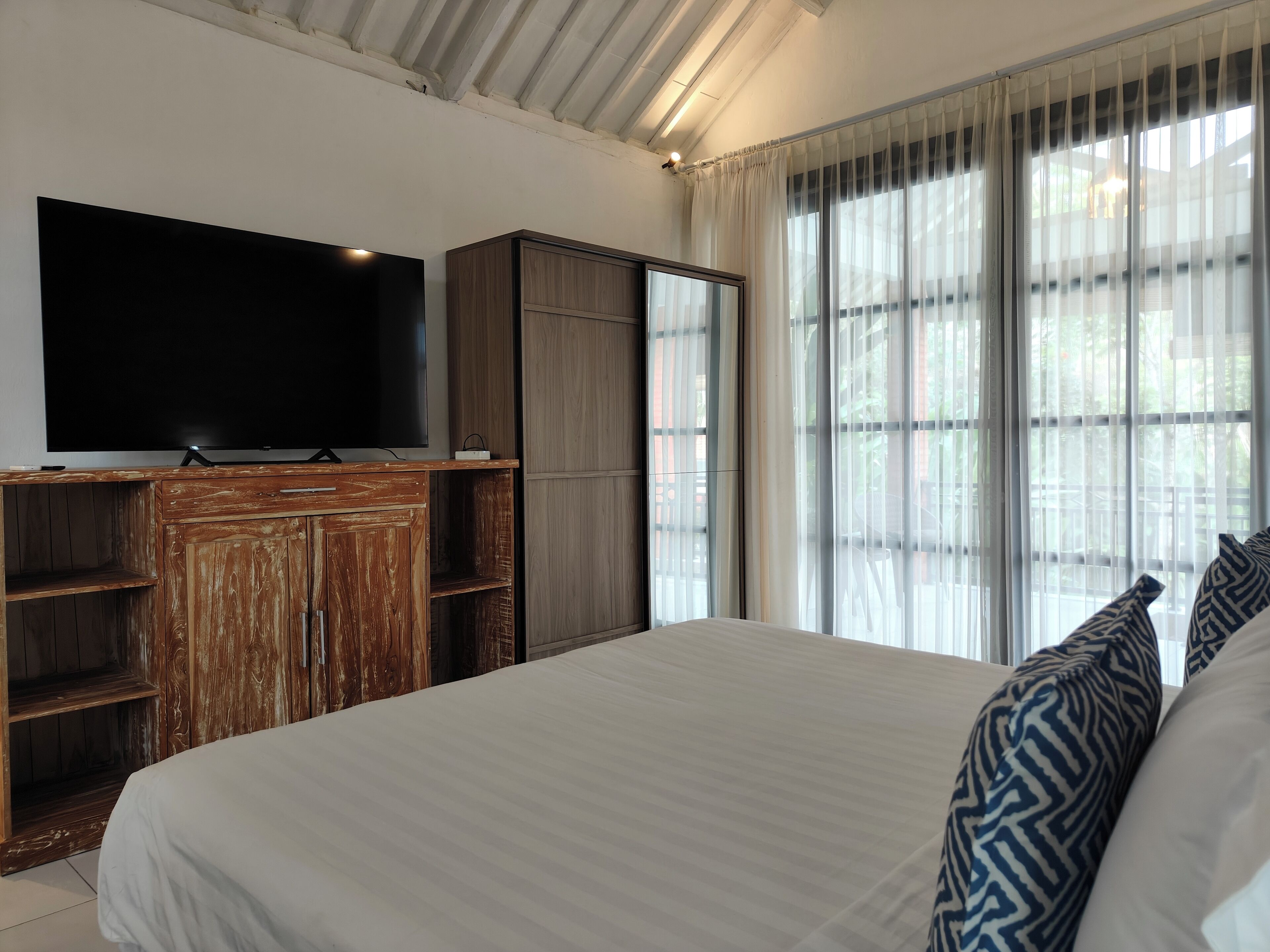 Quarto com cama grande, guarda-roupa, televisão fixada na parede e janela com cortinas leves.