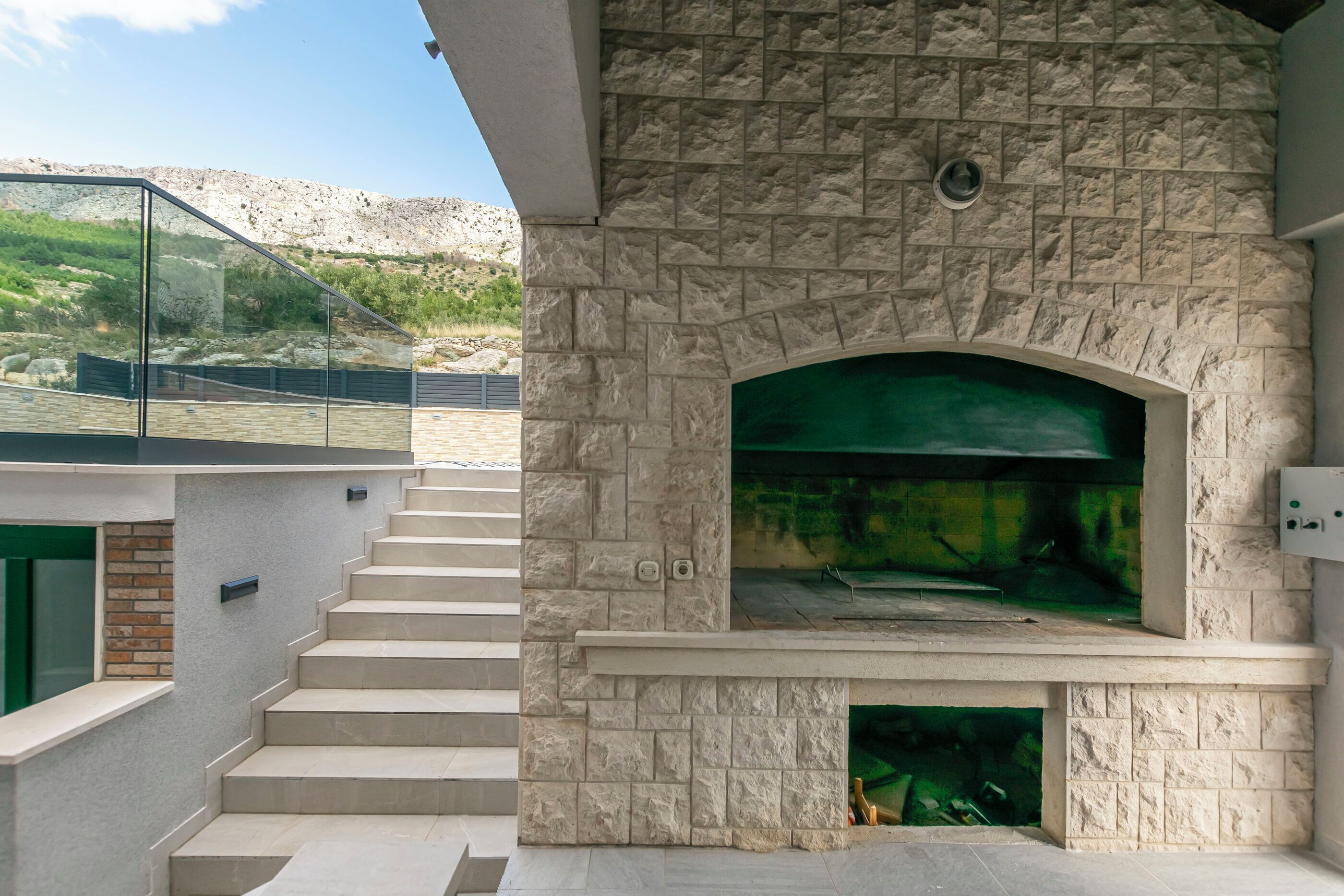 Lareira exterior moderna com fachada de pedra, estrutura de vidro e vista para uma paisagem montanhosa.