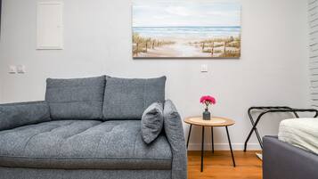 Um quarto com um sofá cinzento, uma mesa redonda pequena com um vaso de flores, e uma pintura de uma praia na parede.